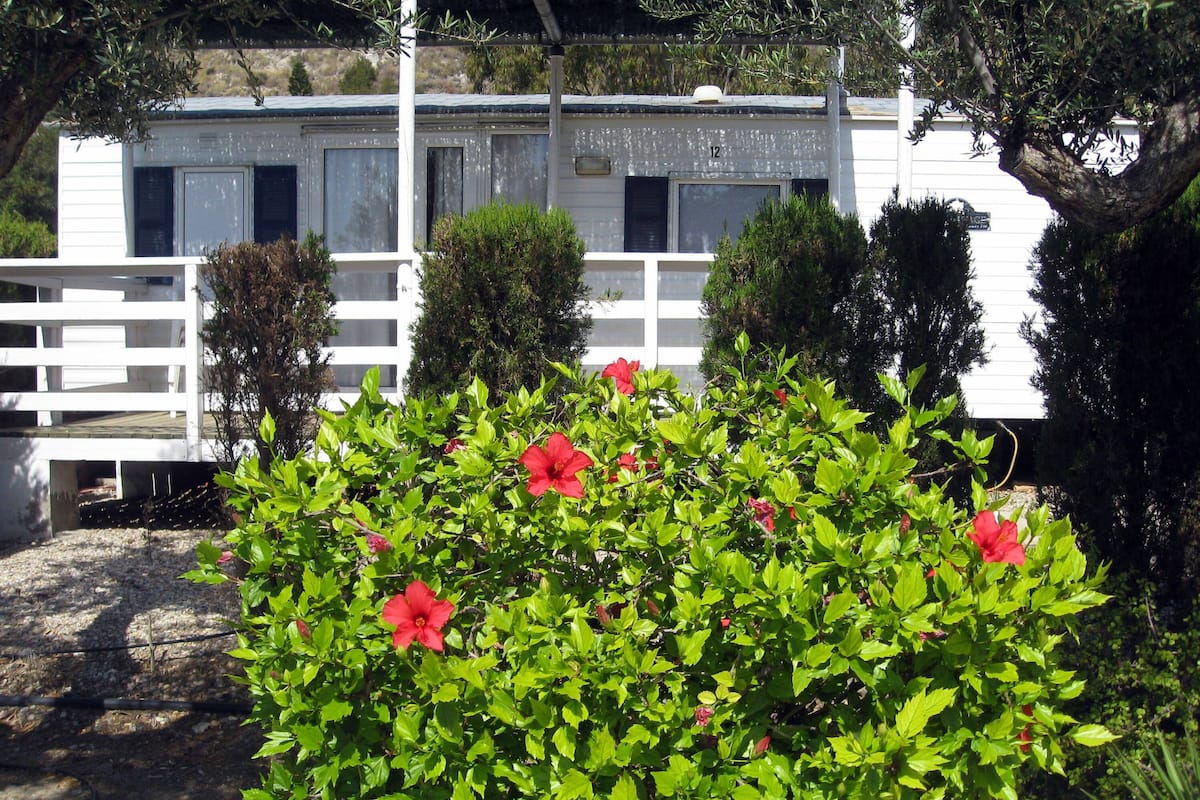 Uma casa móvel branca com um quintal cercado, rodeada por folhagem verde e flores vermelhas.