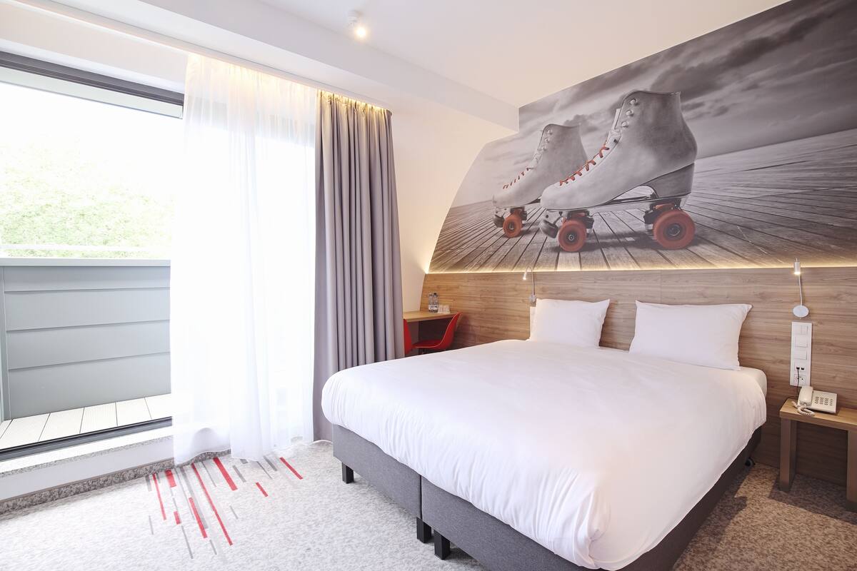 Quarto de hotel moderno com uma cama grande, cabeceira de madeira, mesa de cabeceira com telefone e janela com cortinas.