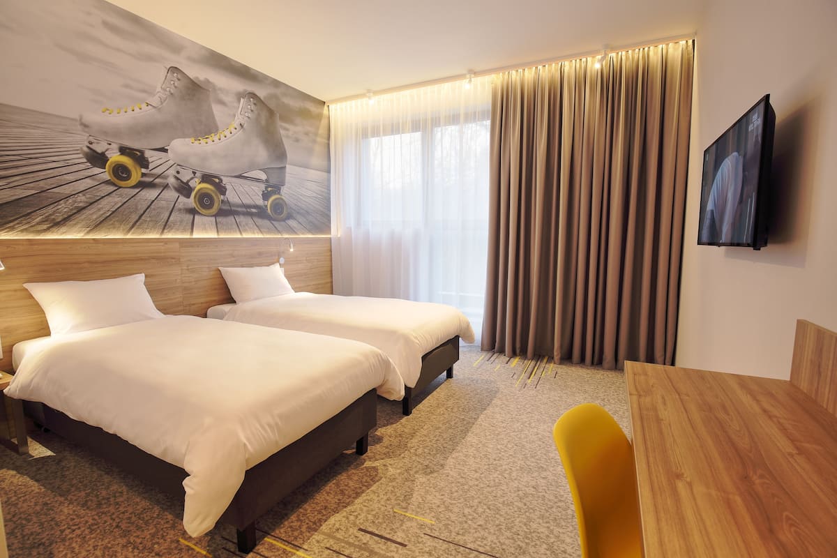 Quarto de hotel com duas camas, cabeceira de madeira, uma televisão grande e uma janela com cortinas.