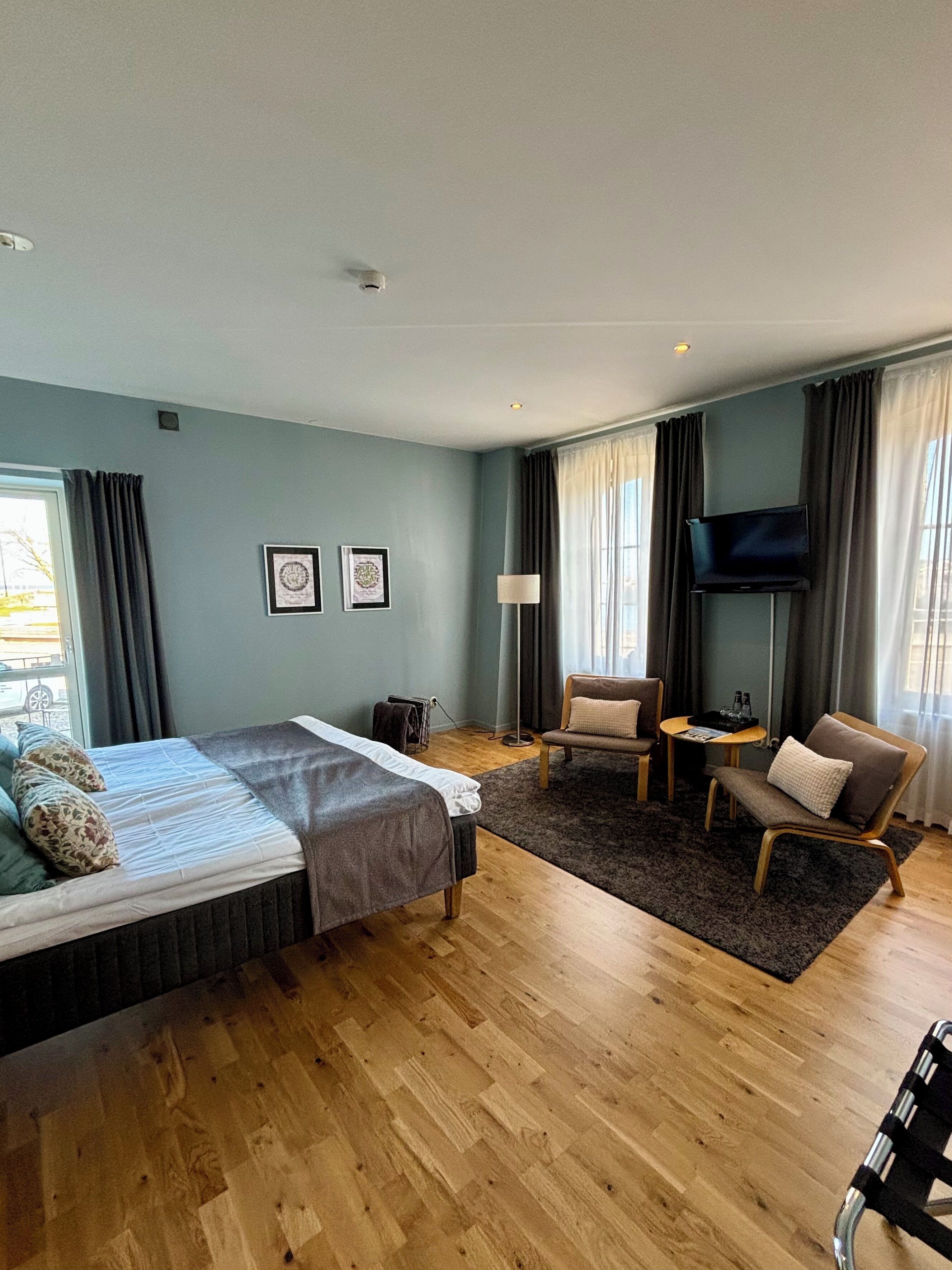 Quarto de hotel com cama, televisão, cadeira, mesa pequena e janela com cortinas.