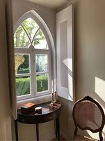 Um quarto com uma grande janela em arco, uma pequena mesa de madeira com uma vela e uma cadeira de vime.