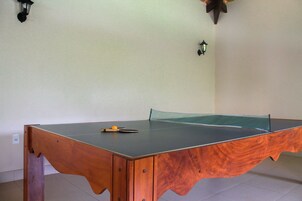 Uma mesa de pingue-pongue com estrutura de madeira e superfície de jogo verde, colocada em um espaço com paredes brancas e duas luzes fixadas na parede.
