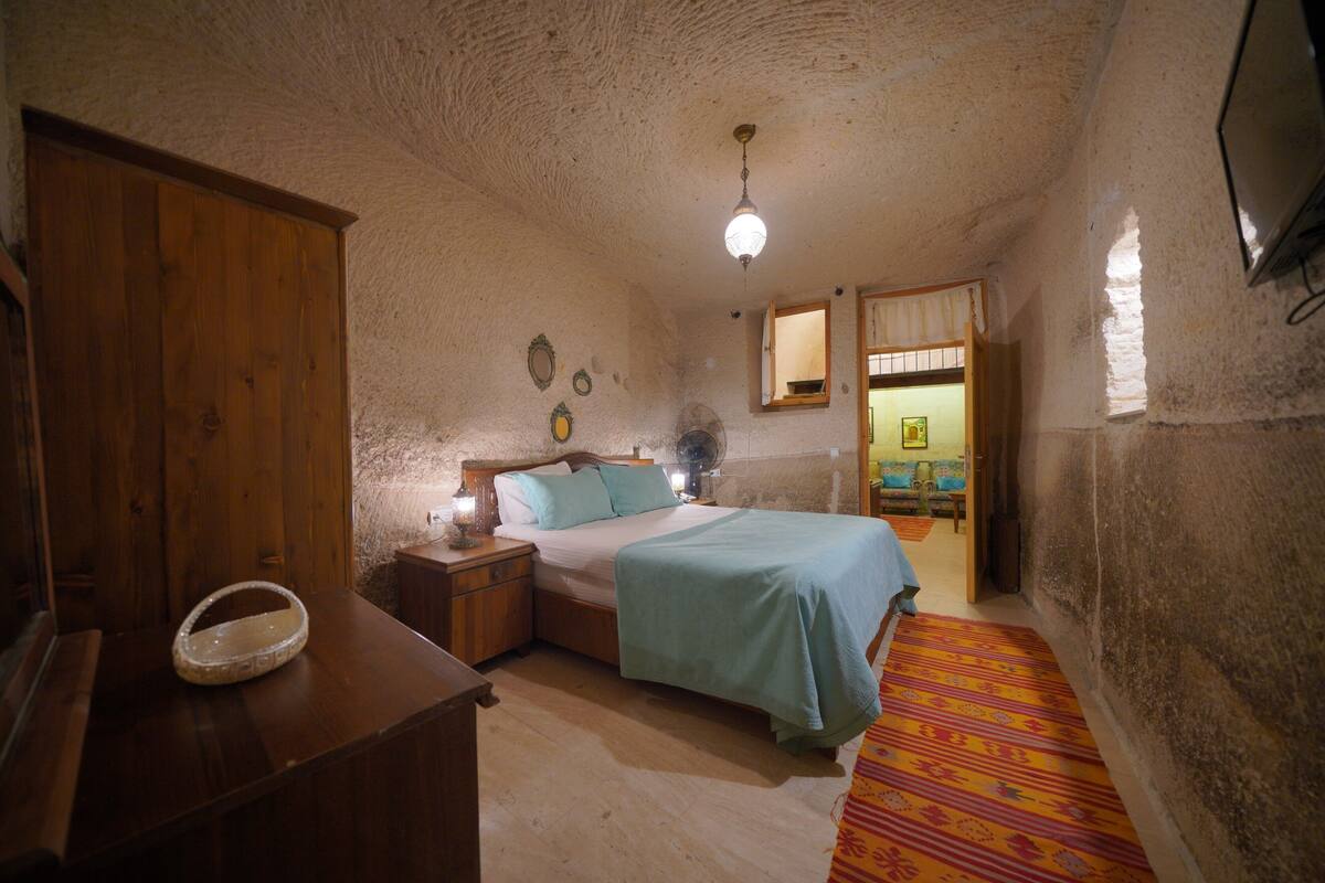 Um quarto com um guarda-roupa de madeira, uma mesa de cabeceira com uma luminária, uma cama com colcha azul, um tapete estampado e uma pequena janela com cortinas.