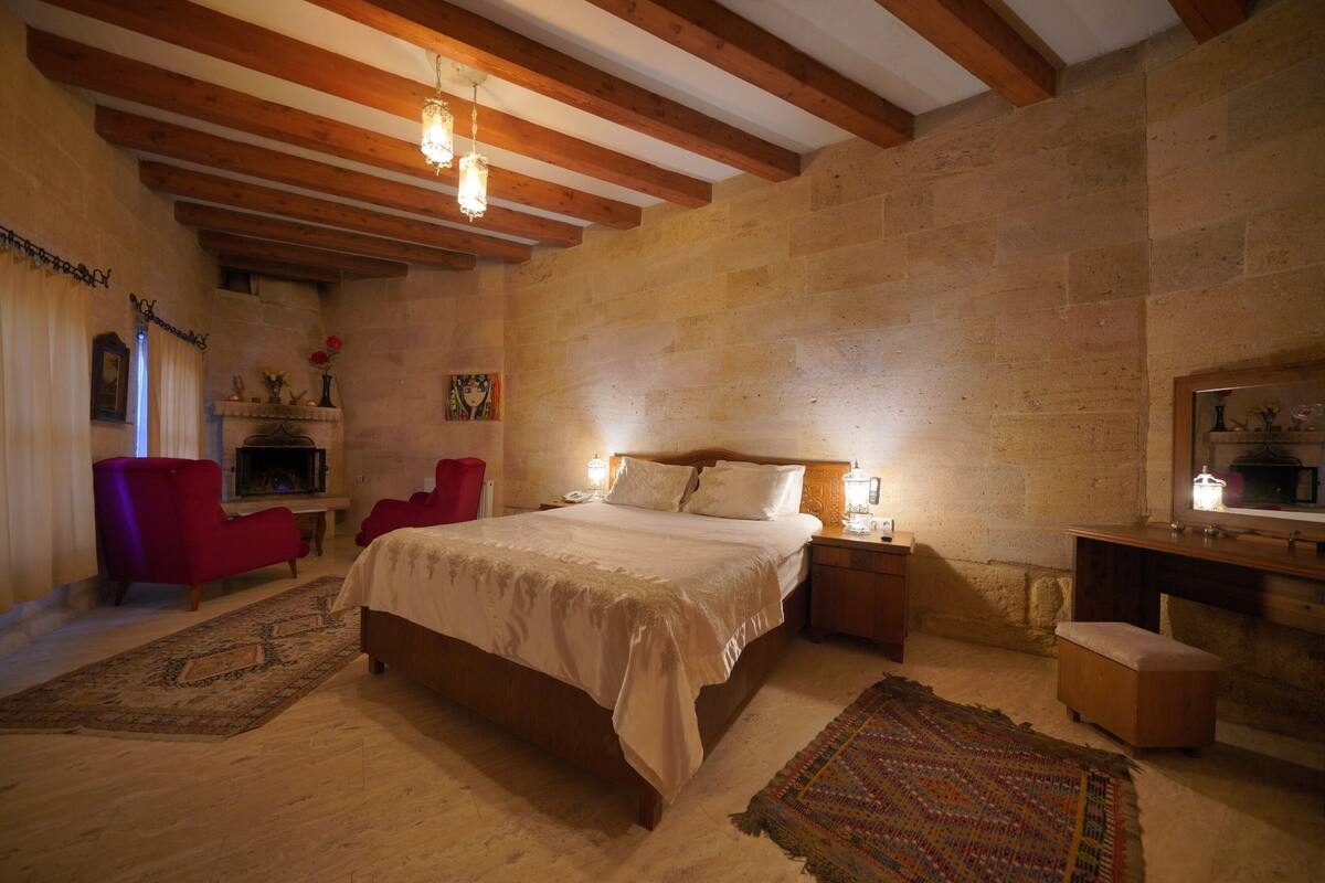Quarto com cama grande, vigas de madeira, paredes de pedra, poltrona vermelha e pequena mesa com espelho.