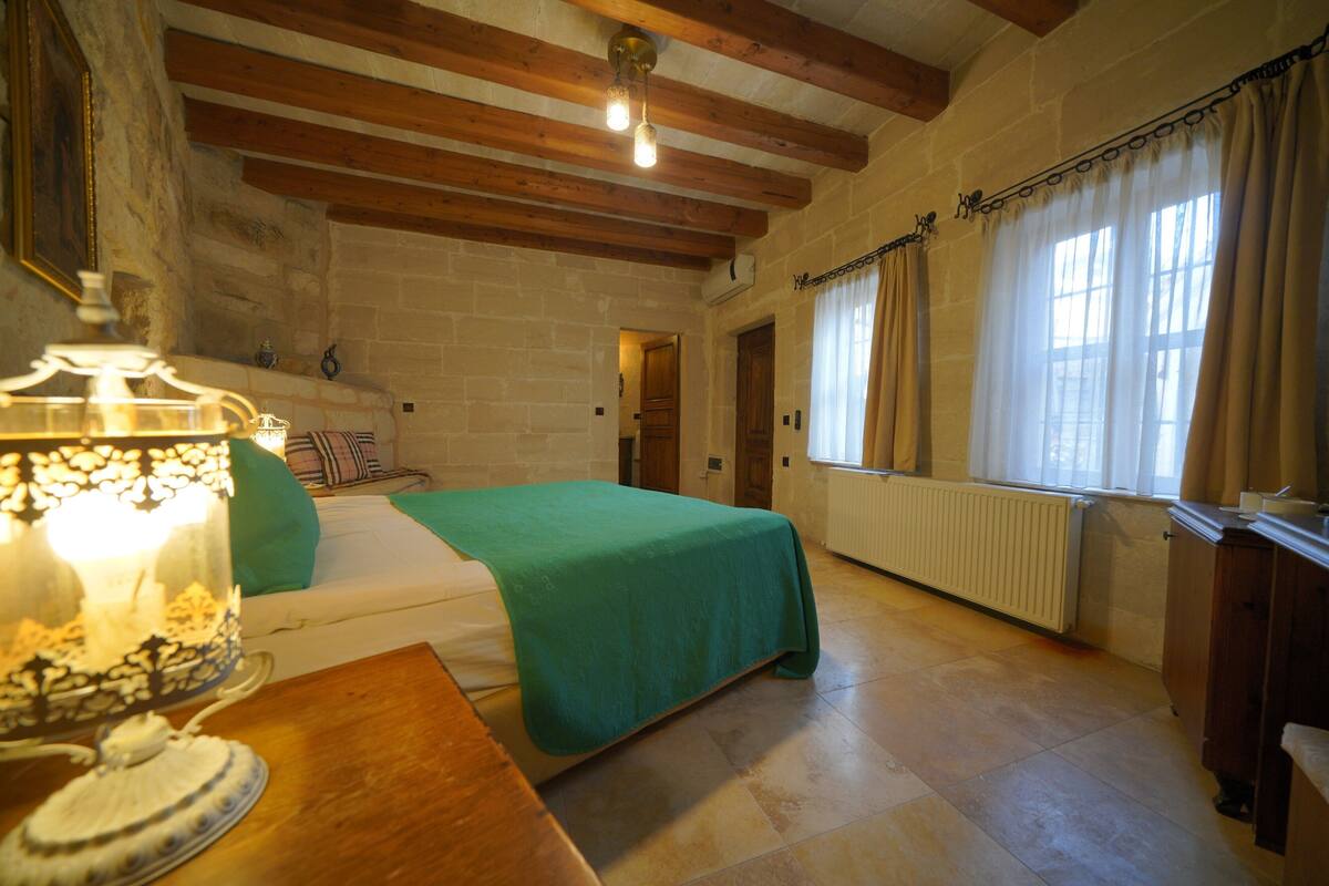 Quarto com cama, mesa com luminária, janela com cortinas e radiador.