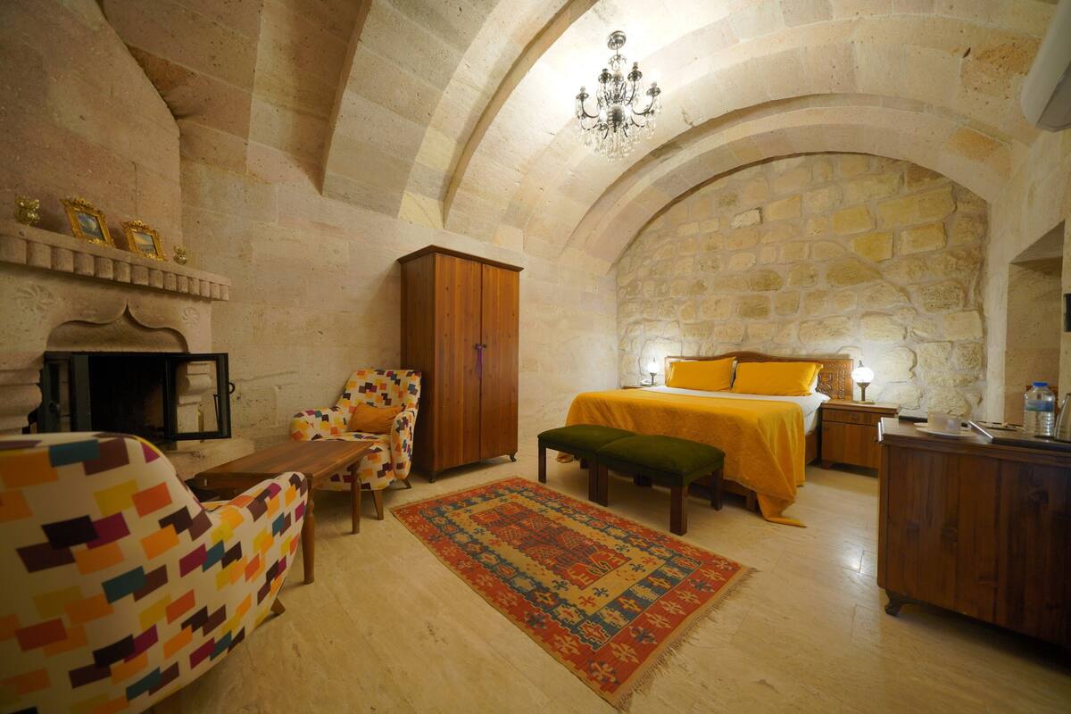 Quarto com parede de pedra, cama com roupa de cama amarela, guarda-roupa de madeira, cadeira de madeira, tapete colorido e um candelabro.