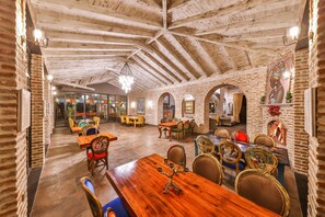 Uma sala de jantar ampla, com pé direito alto e paredes de tijolo aparente, com várias mesas e cadeiras.