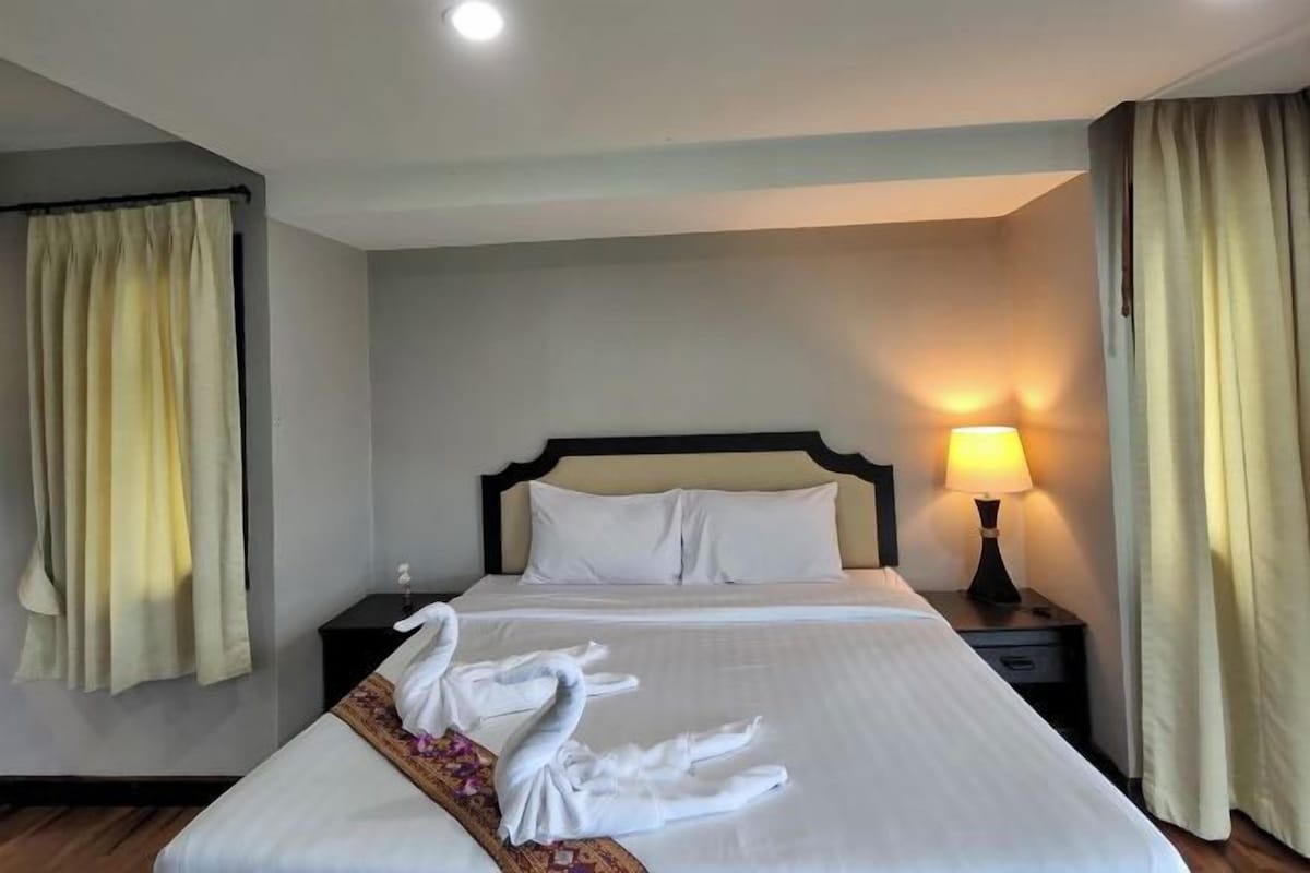 Quarto de hotel com uma cama grande, duas mesas de cabeceira, uma luminária e cortinas.
