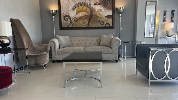 Uma sala de estar moderna com um sofá bege, uma mesa de centro de vidro e um banquinho vermelho. Há um quadro abstrato grande na parede e duas luminárias de chão.