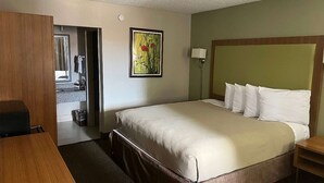 Quarto de hotel com cama, escrivaninha, cadeira, luminária e um quadro na parede.