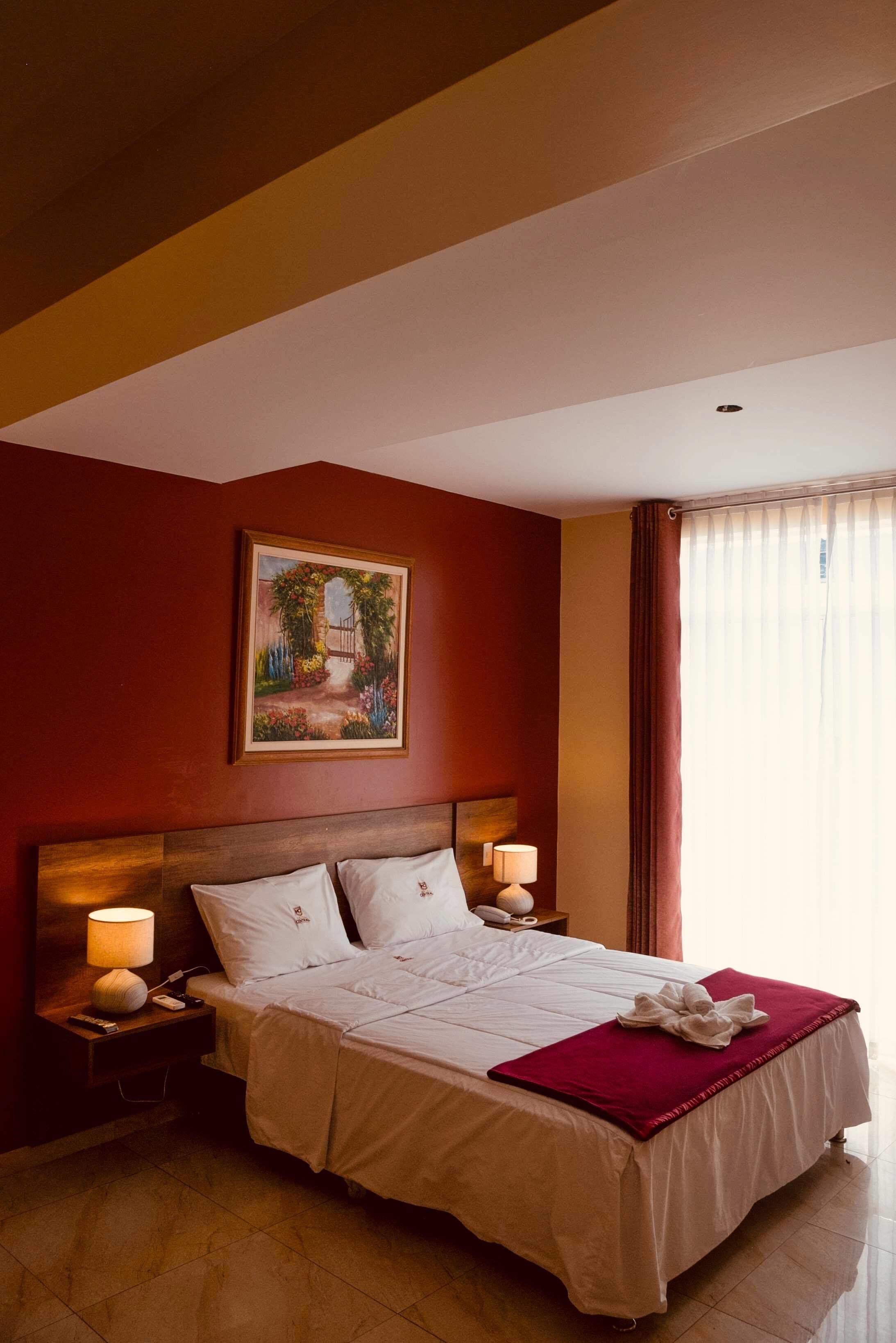 Quarto de hotel com uma cama grande, mesa de cabeceira com abajur, pintura na parede e janela com cortinas.