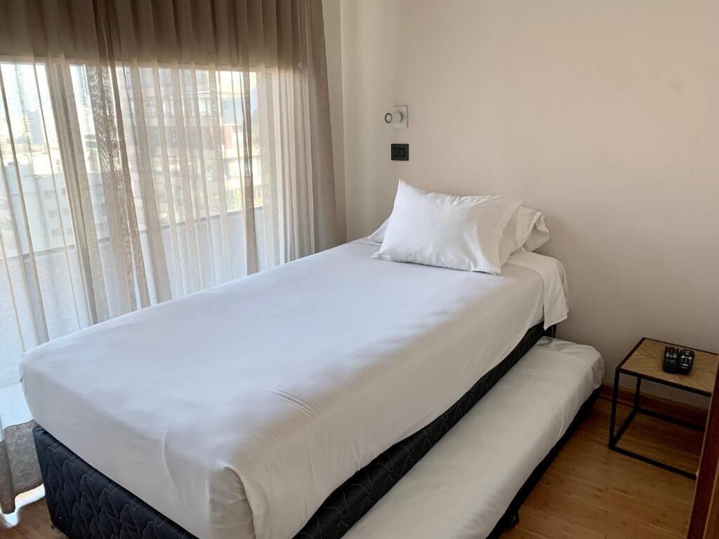 Cama bem arrumada com lençóis brancos em quarto com piso de madeira e uma mesa pequena com alguns objetos.