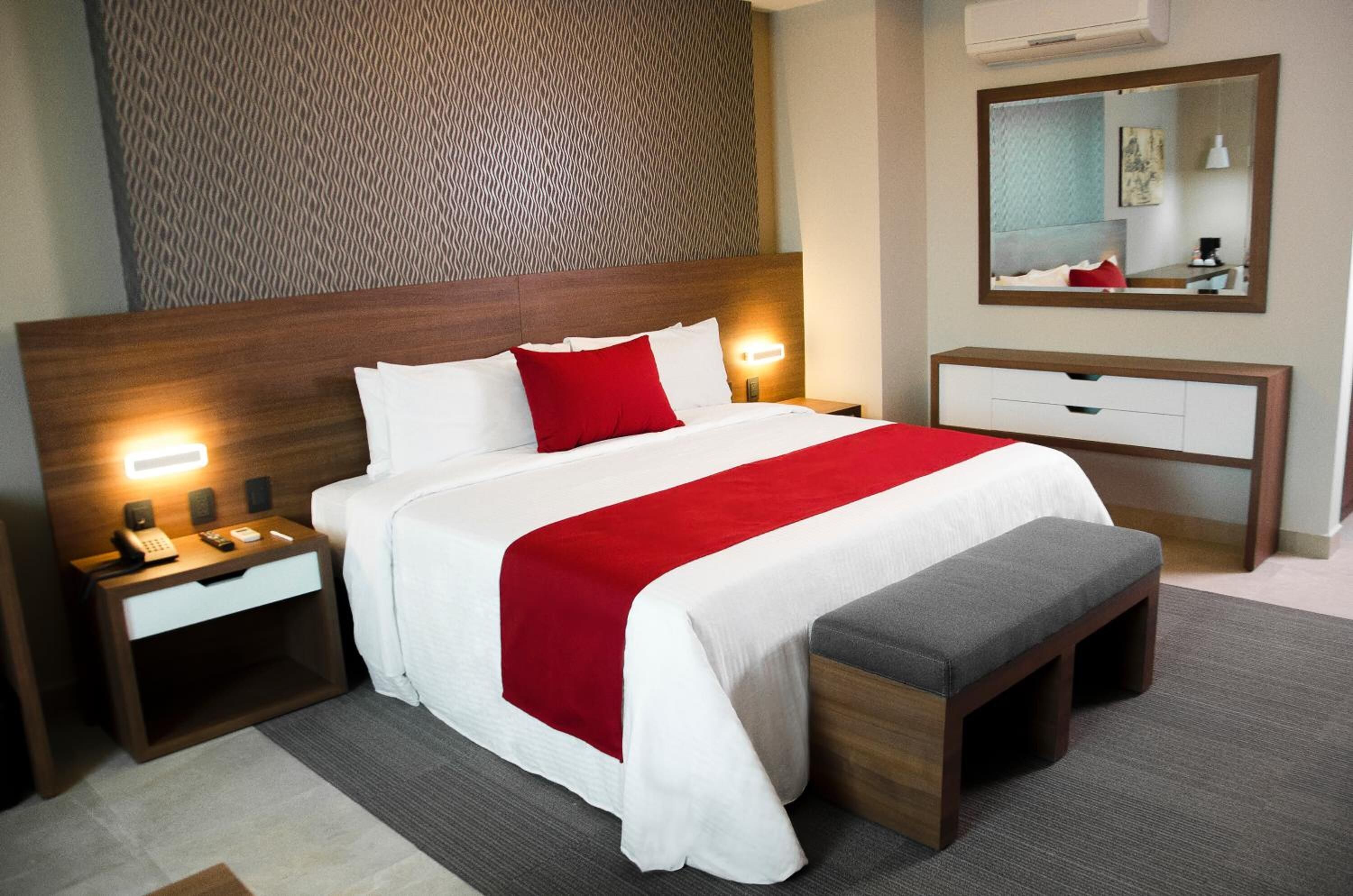 Quarto de hotel com uma cama grande, mesas de cabeceira, um banco, cômoda e um espelho.
