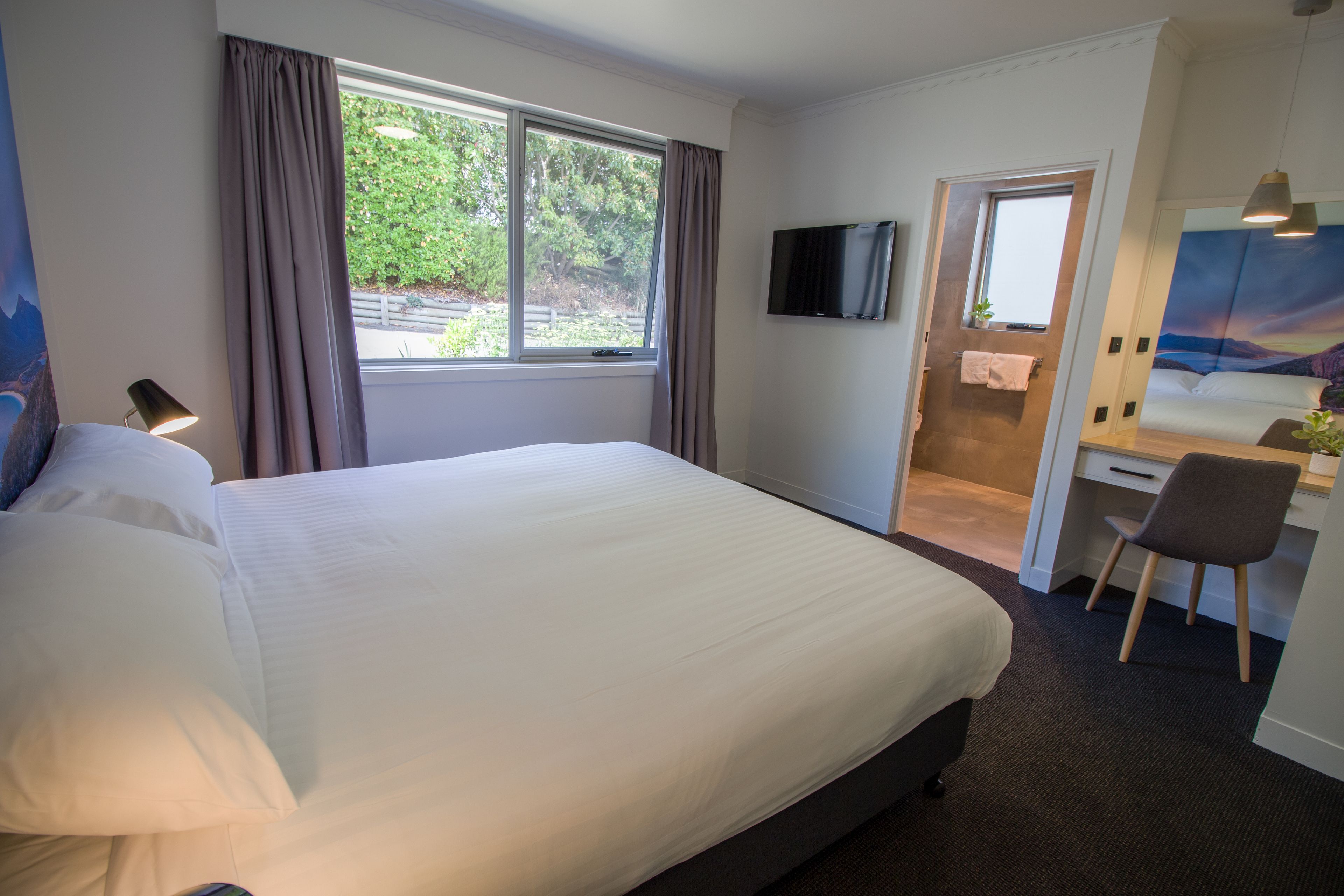Quarto de hotel com uma cama grande, uma mesa com cadeira, uma televisão e uma janela com cortinas.