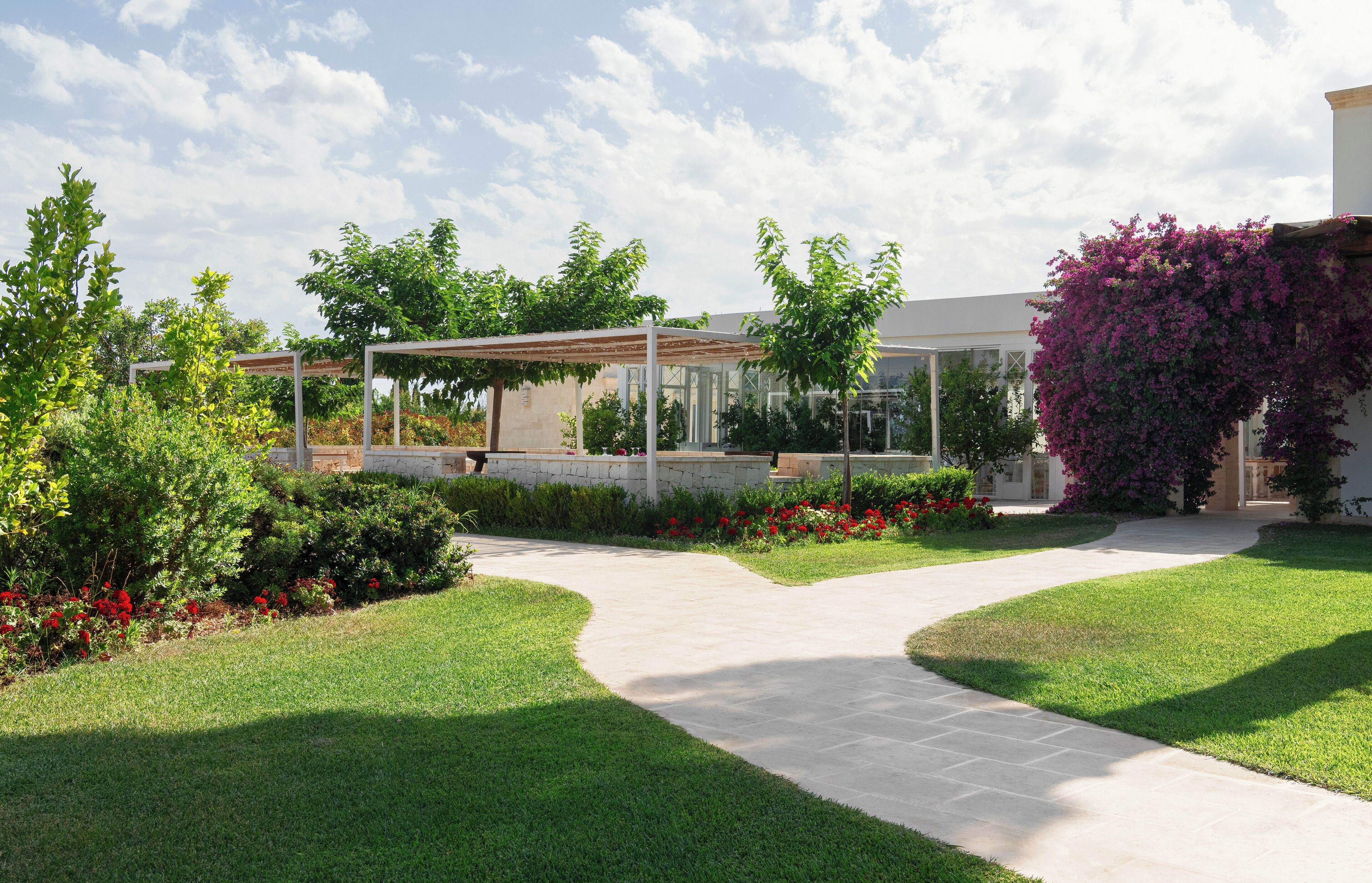 Um jardim com um caminho pavimentado, uma pérgula e um edifício com fachada de vidro.
