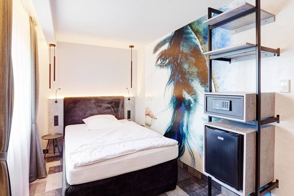 Quarto de hotel moderno com cama, pequena escrivaninha, frigorífico e uma grande pintura abstrata na parede.