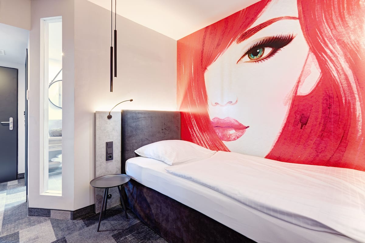 Quarto de hotel moderno com uma cama grande, uma mesinha de cabeceira pequena, um lustre moderno e um grande mural na parede.