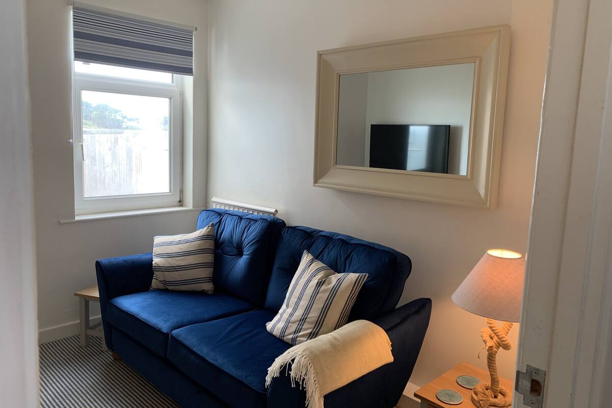 Quarto com sofá azul, almofadas listradas, uma mesinha com abajur e um espelho na parede.
