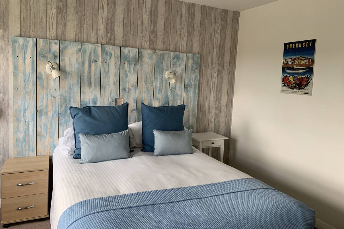 Um quarto com cabeceira de madeira, duas mesas de cabeceira, uma cama com roupa de cama azul e um cartaz na parede.