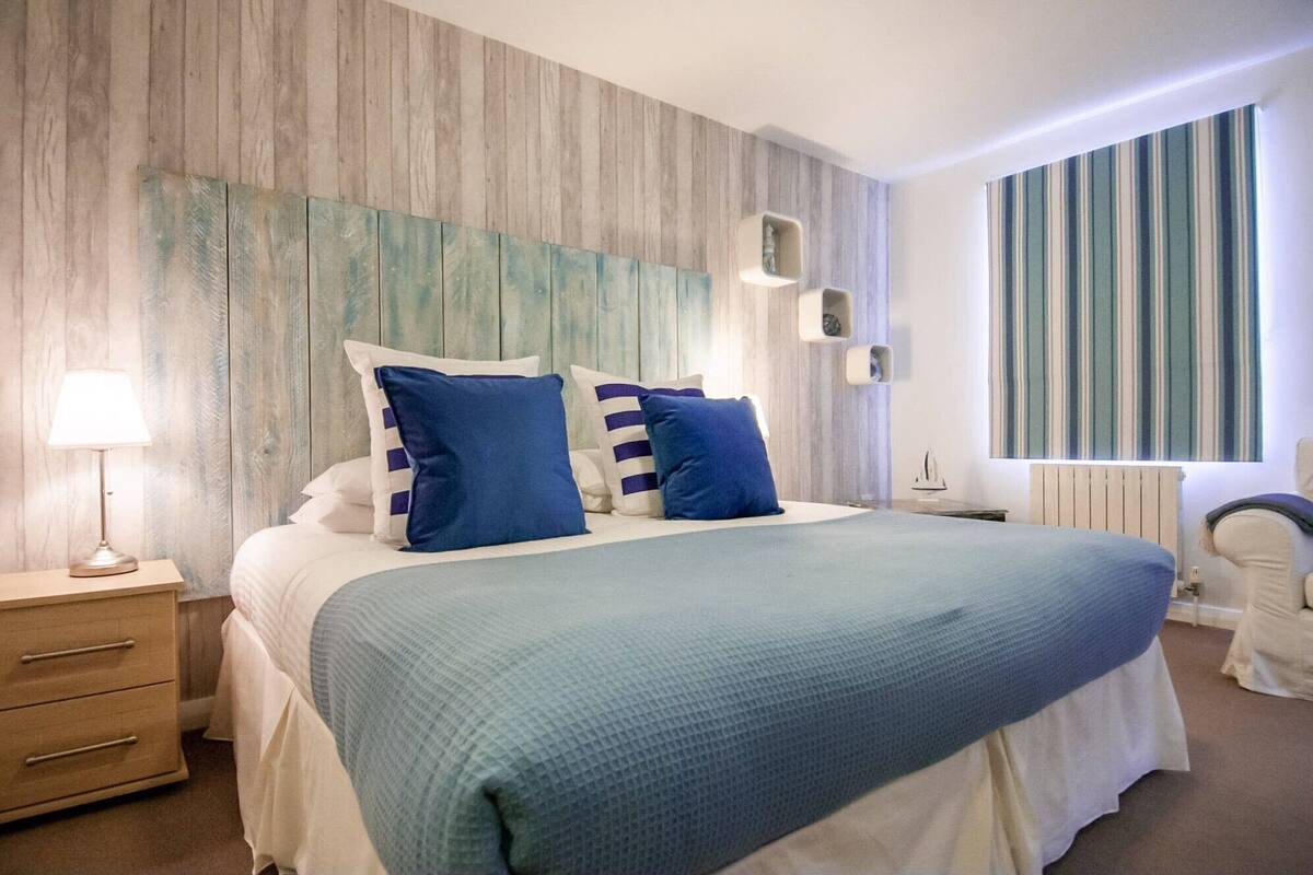 Quarto com cama, mesa de cabeceira, luminária, cômoda e janela com cortinas listradas.
