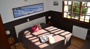Quarto com uma cama grande, mesa de cabeceira, abajur, janela com vista para árvores e um quadro na parede.