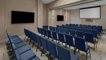 Uma sala de conferência com fileiras de cadeiras azuis, uma tela grande e um projetor.