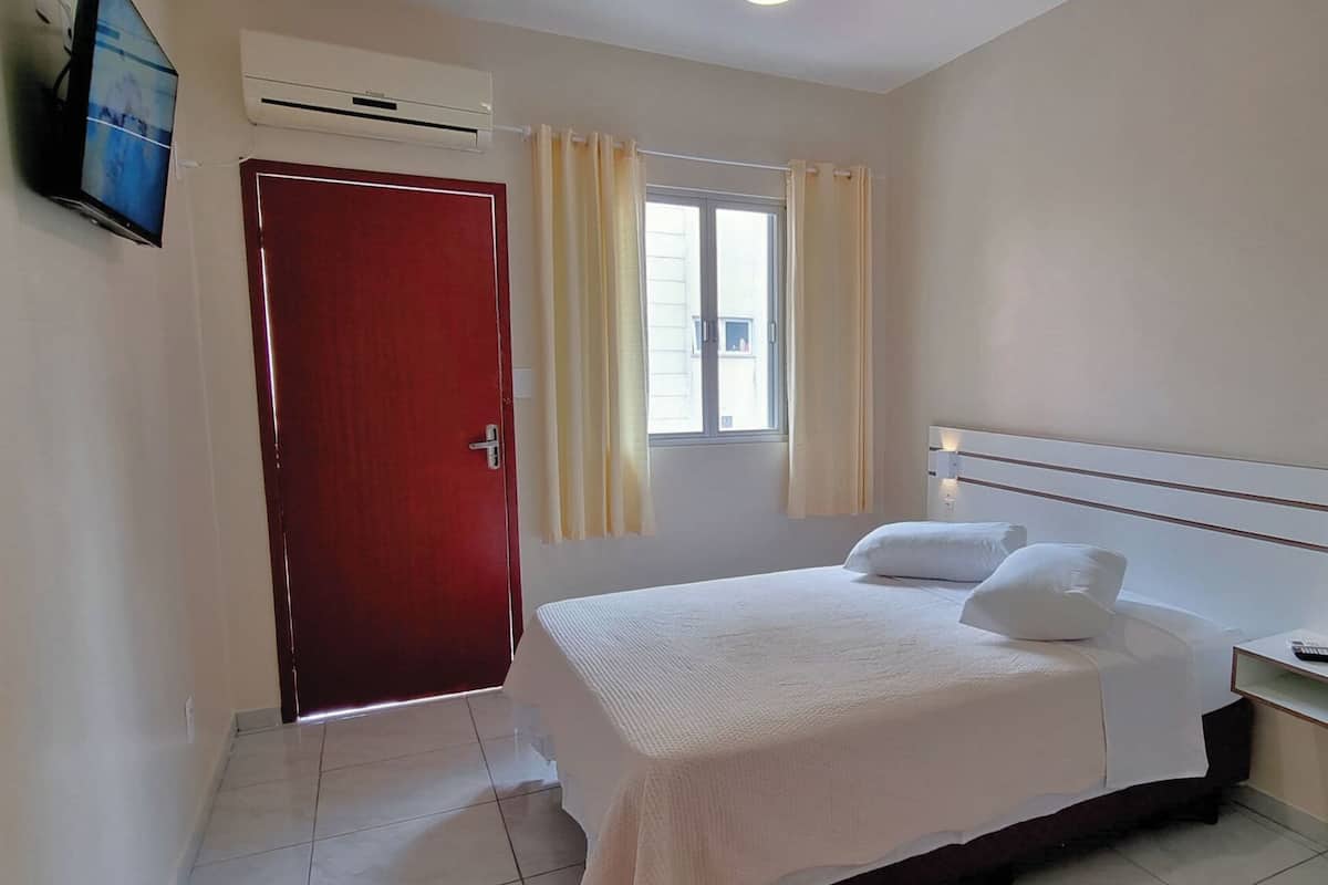 Quarto de hotel com uma cama, mesa de cabeceira, televisão na parede, ar condicionado, janela com cortinas e uma porta vermelha.