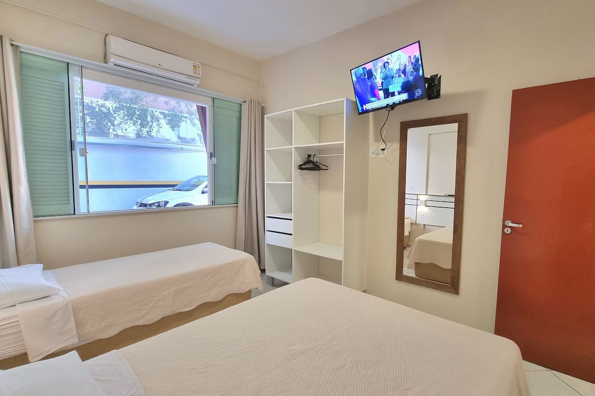 Quarto de hotel com duas camas de solteiro, televisão fixada na parede, armário e janela com cortinas.