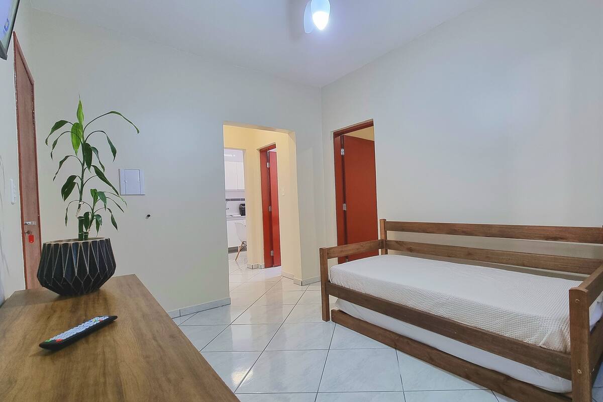 Quarto com sofá de madeira, uma planta em vaso e controle remoto sobre uma mesa de madeira.
