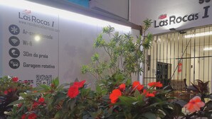 Um edifício com uma placa indicando 'Las Rocas I' e comodidades listadas, incluindo Wi-Fi, proximidade à praia e uma garagem rotativa.
