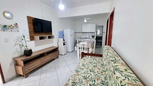 Uma sala de estar moderna com uma TV de tela plana, unidade de entretenimento de madeira e um sofá. Uma cozinha é visível ao fundo, com uma área de refeições.