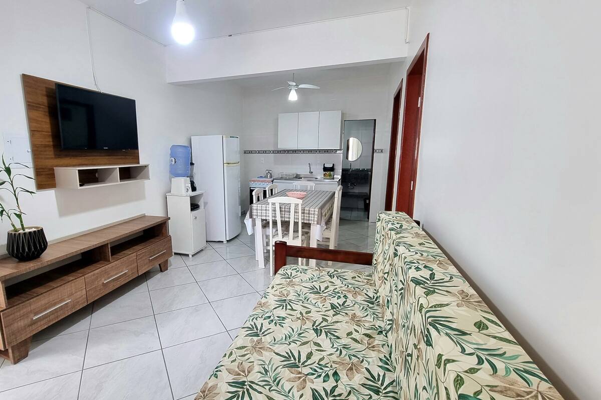 Uma sala de estar moderna com uma TV de tela plana, unidade de entretenimento de madeira e um sofá. Uma cozinha é visível ao fundo, com uma área de refeições.