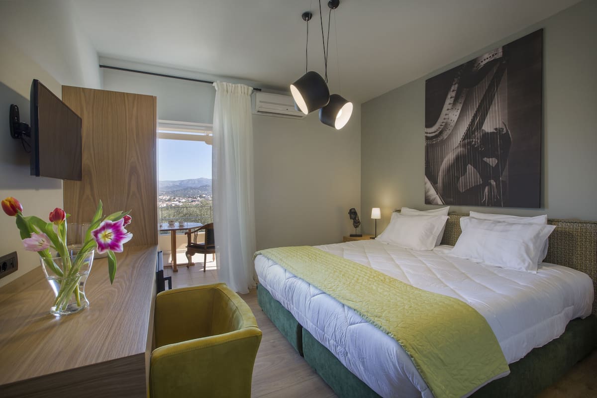 Quarto de hotel com cama, escrivaninha, cadeira, vaso com flores e vista para a cidade.