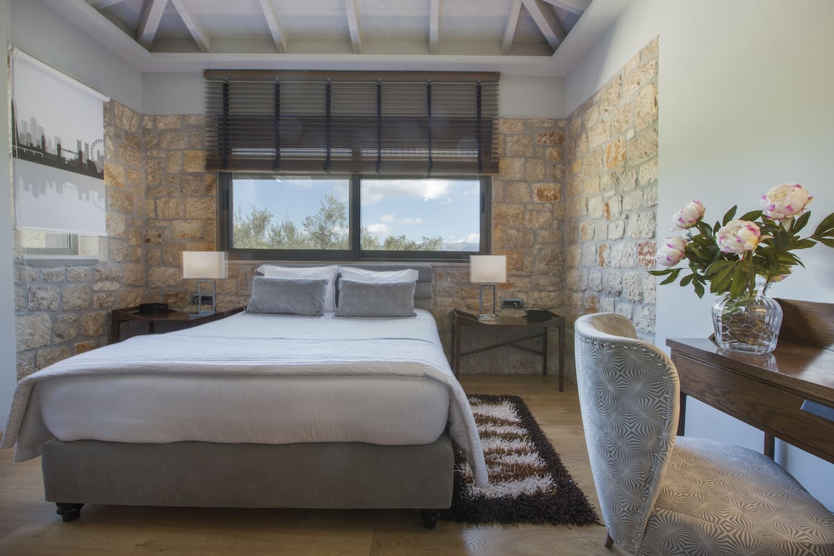 Quarto com parede de pedra, uma cama grande, mesa de cabeceira, abajur, janela com persianas e um vaso com flores.