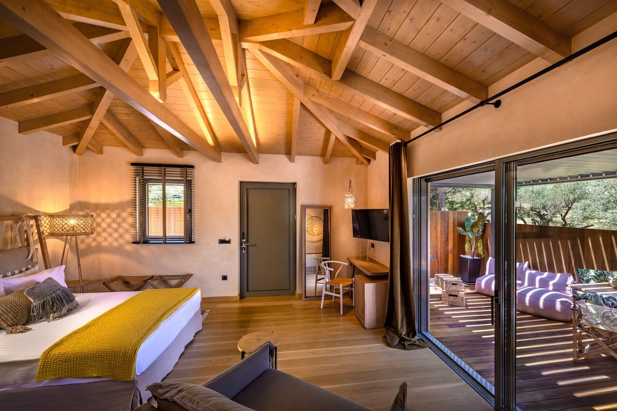 Quarto espaçoso com teto de madeira, cama com cobertor amarelo, escrivaninha com cadeira e vista para uma área de estar externa.