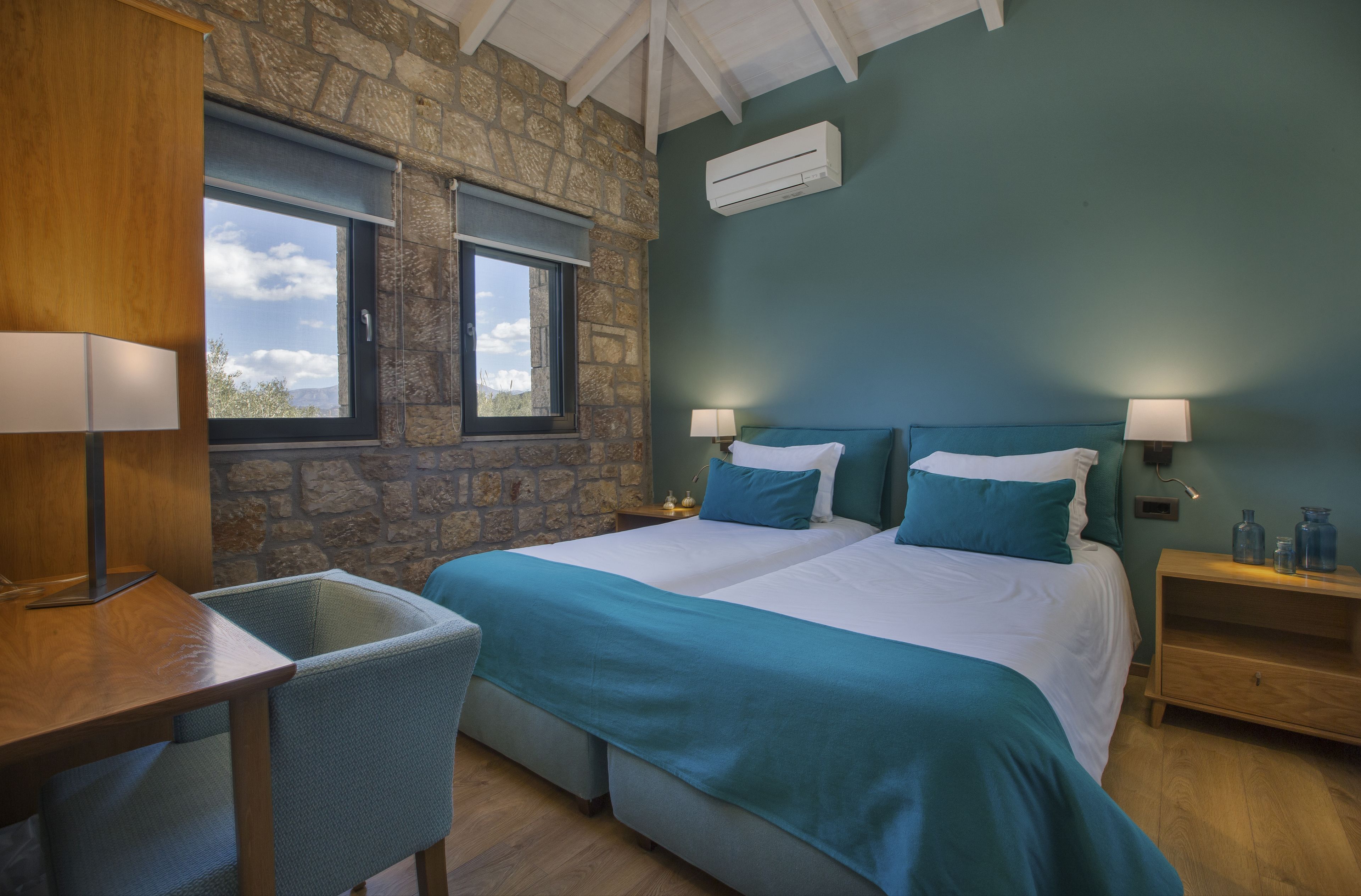 Quarto com parede de pedra, cama com roupa de cama azul, mesa de cabeceira de madeira e escrivaninha com cadeira.