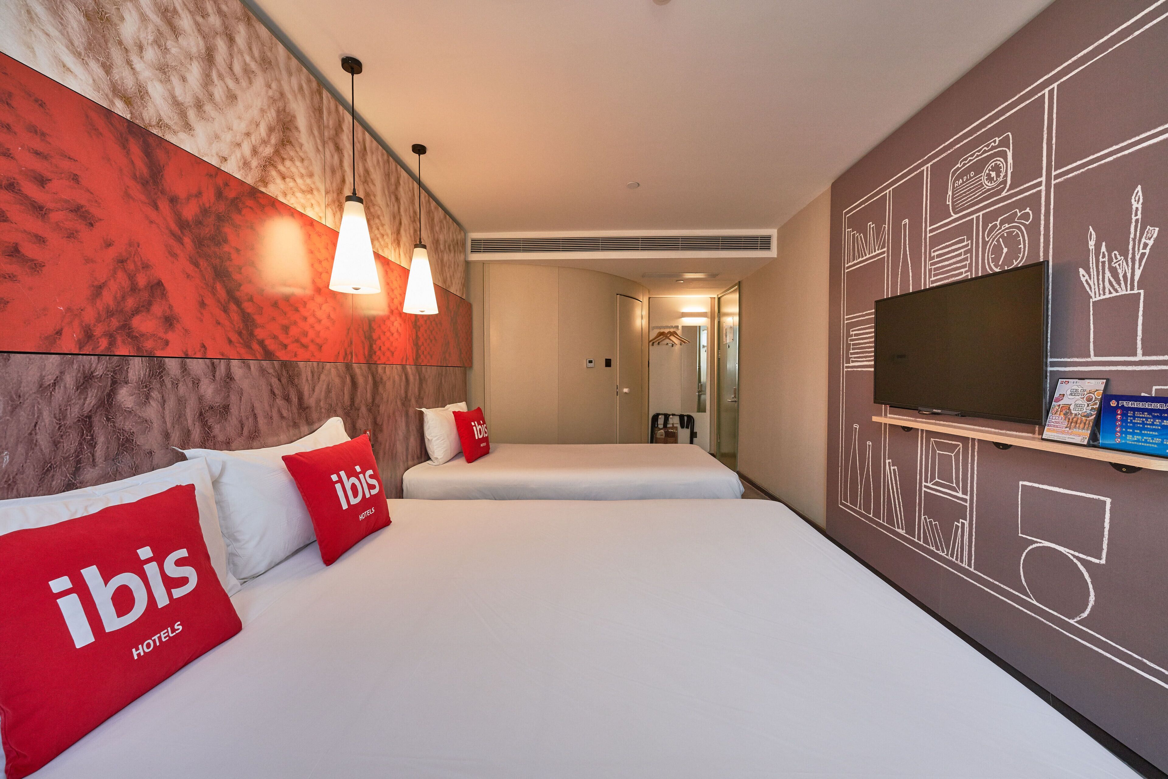Quarto de hotel com duas camas, almofadas vermelhas e brancas, uma televisão montada e uma parede com desenhos a linha em branco.