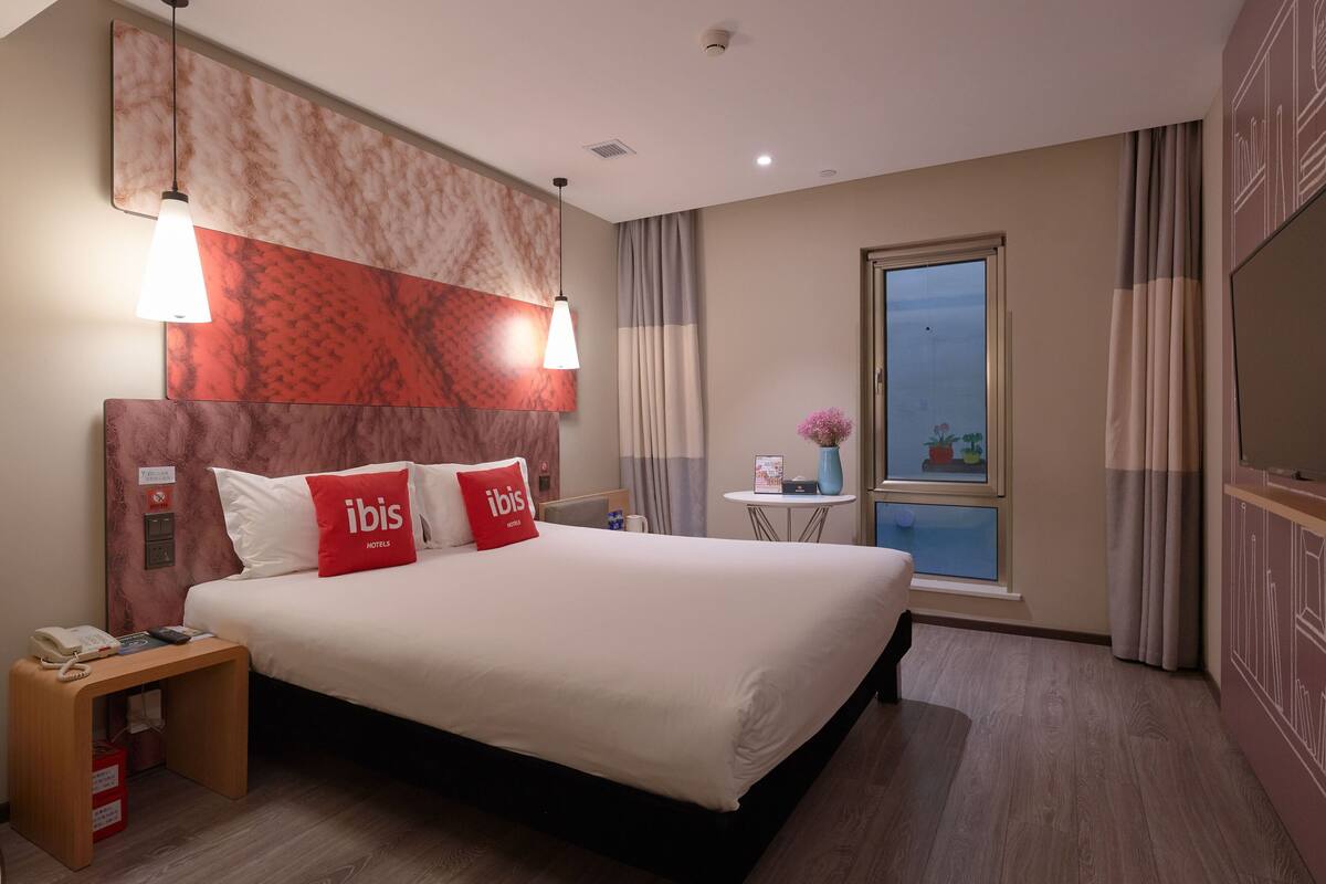 Quarto de hotel com uma cama grande, duas luminárias de cabeceira, uma mesa de cabeceira com telefone, uma mesinha com um vaso e uma janela com vista.