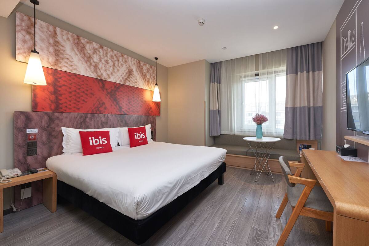Quarto de hotel com cama grande, mesas de cabeceira, escrivaninha e janela com cortinas.