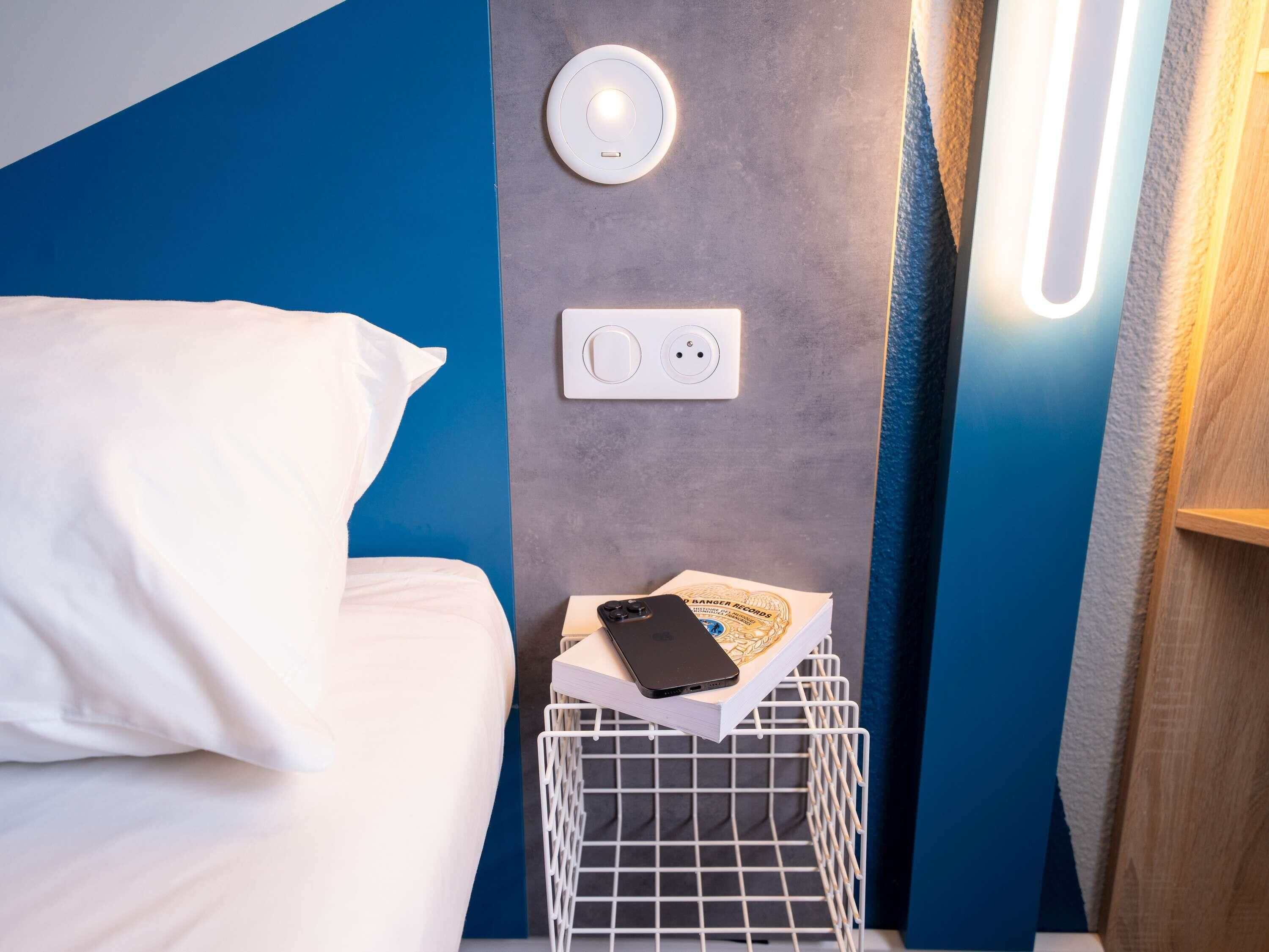 Quarto de hotel com cama, mesa de cabeceira com abajur, tomada e luminária na parede.