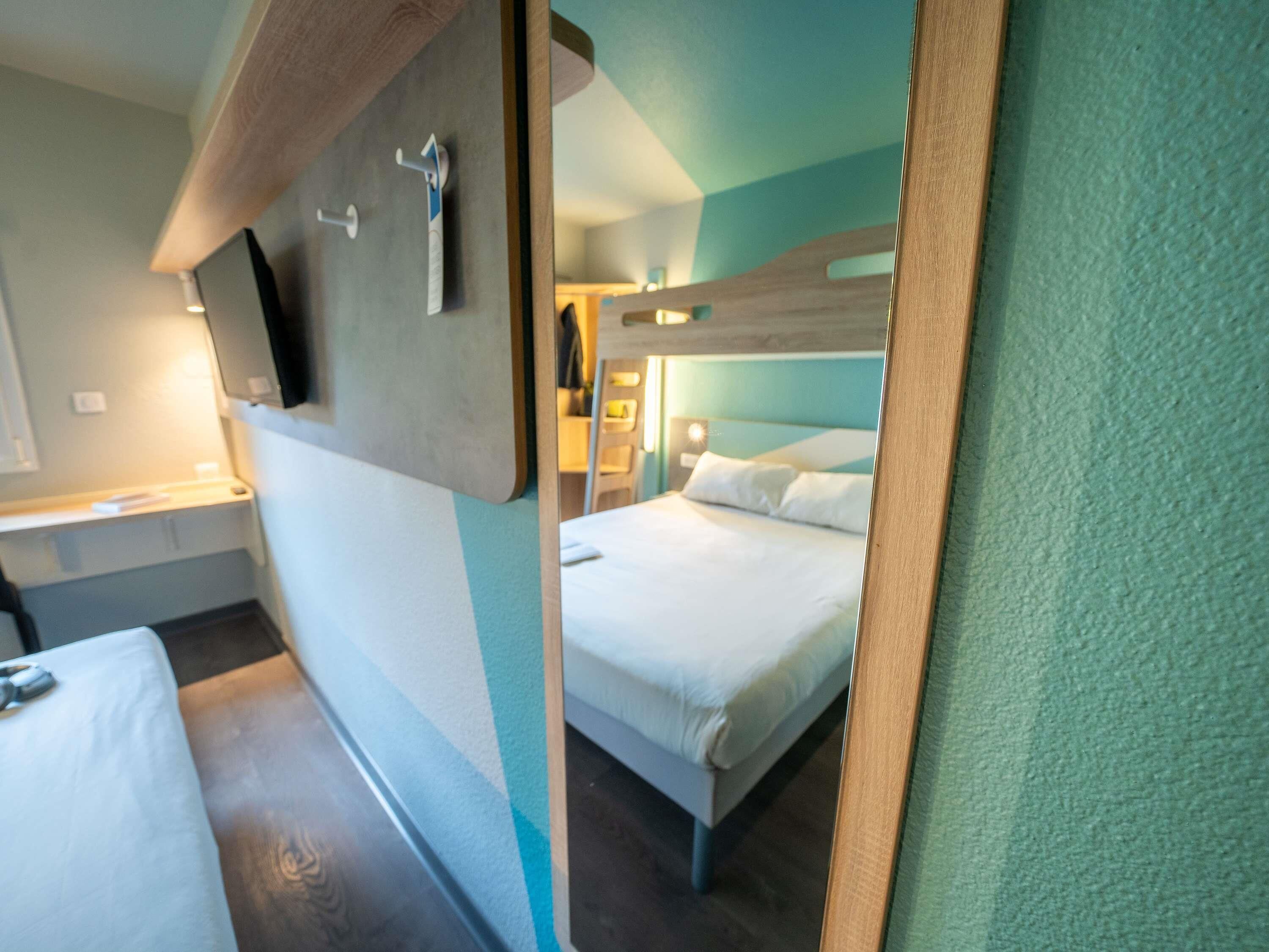 Quarto de hotel com cama beliche, escrivaninha com cadeira e espelho refletindo o quarto.