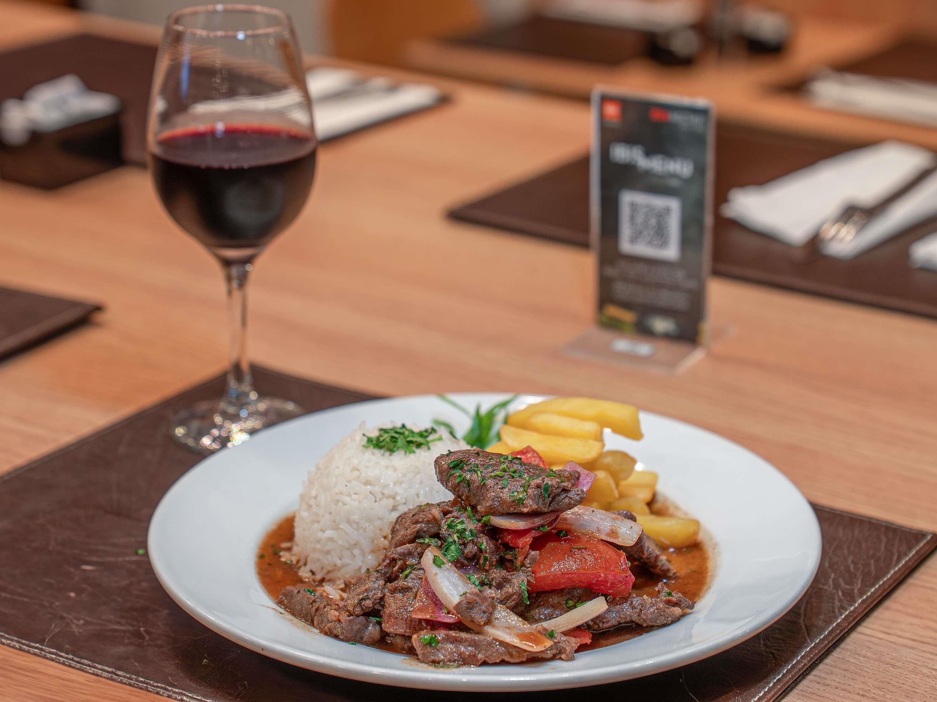 Prato de carne com legumes e arroz, um copo de vinho tinto e um menu com código QR sobre uma mesa de madeira.