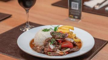Prato de carne com legumes e arroz, um copo de vinho tinto e um menu com código QR sobre uma mesa de madeira.