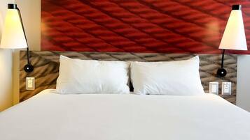 Quarto de hotel com uma cama grande, duas luminárias de cabeceira, um encosto de cama vermelho com textura e uma tomada.