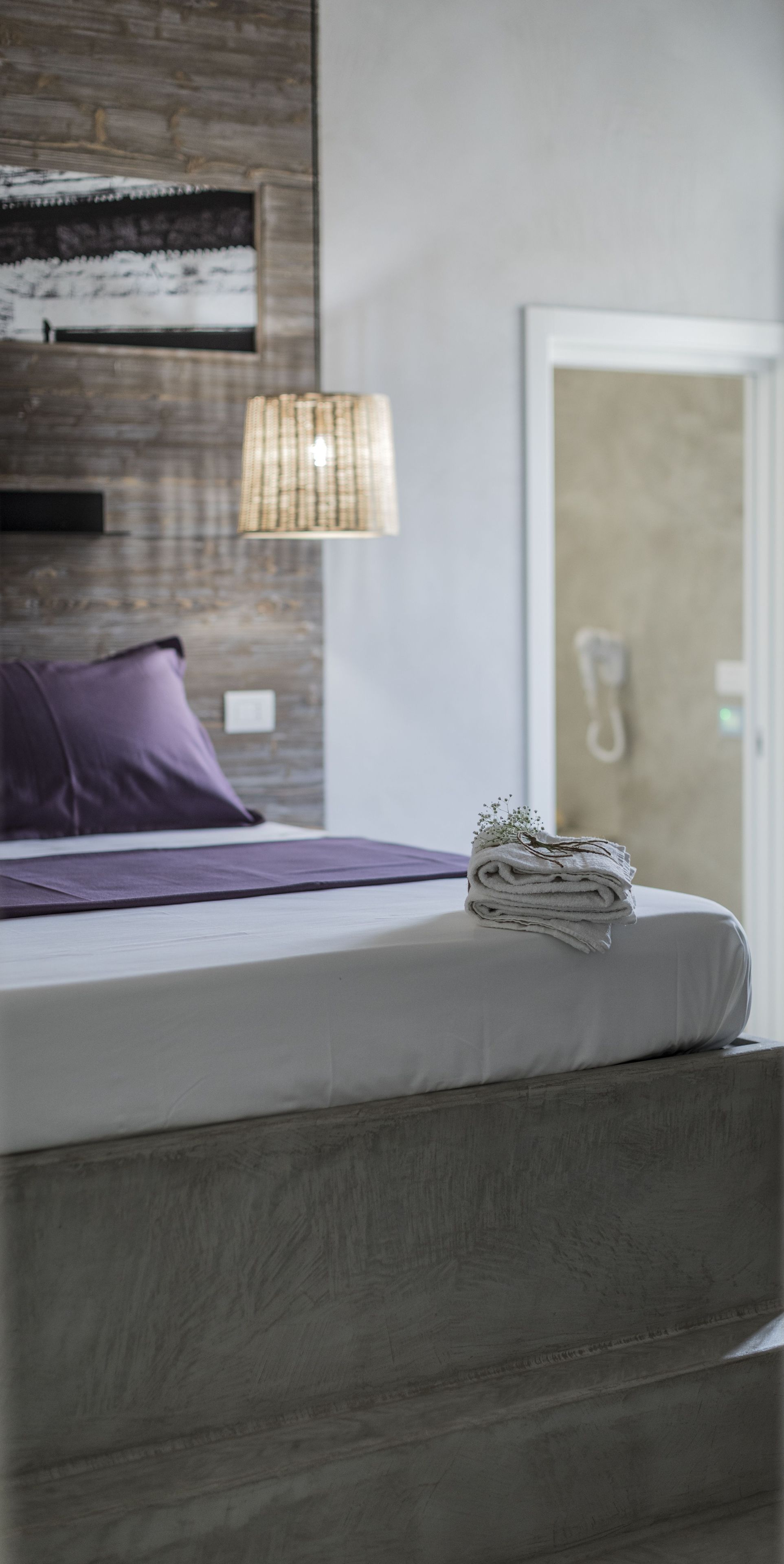 Cama bem arrumada com travesseiro roxo e toalha dobrada em uma cabeceira de madeira.