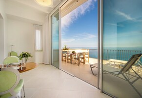 Uma varanda com vista para o mar, uma área de refeições com cadeiras e uma mesa, e uma porta de vidro deslizante.