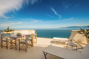 Um terraço com mesa e cadeiras, uma espreguiçadeira e vista para o mar.