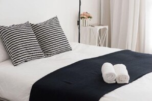 Cama bem arrumada com travesseiro xadrez preto e branco, manta preta e duas toalhas brancas enroladas.