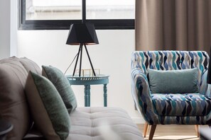Uma sala de estar moderna com um sofá, uma poltrona estampada, uma mesinha de centro com um abajur e vista pela janela.