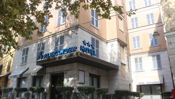 Um hotel com uma placa chamativa com a inscrição "HOTEL ALEXANDER" e fachada moderna com janelas simétricas e entrada central.